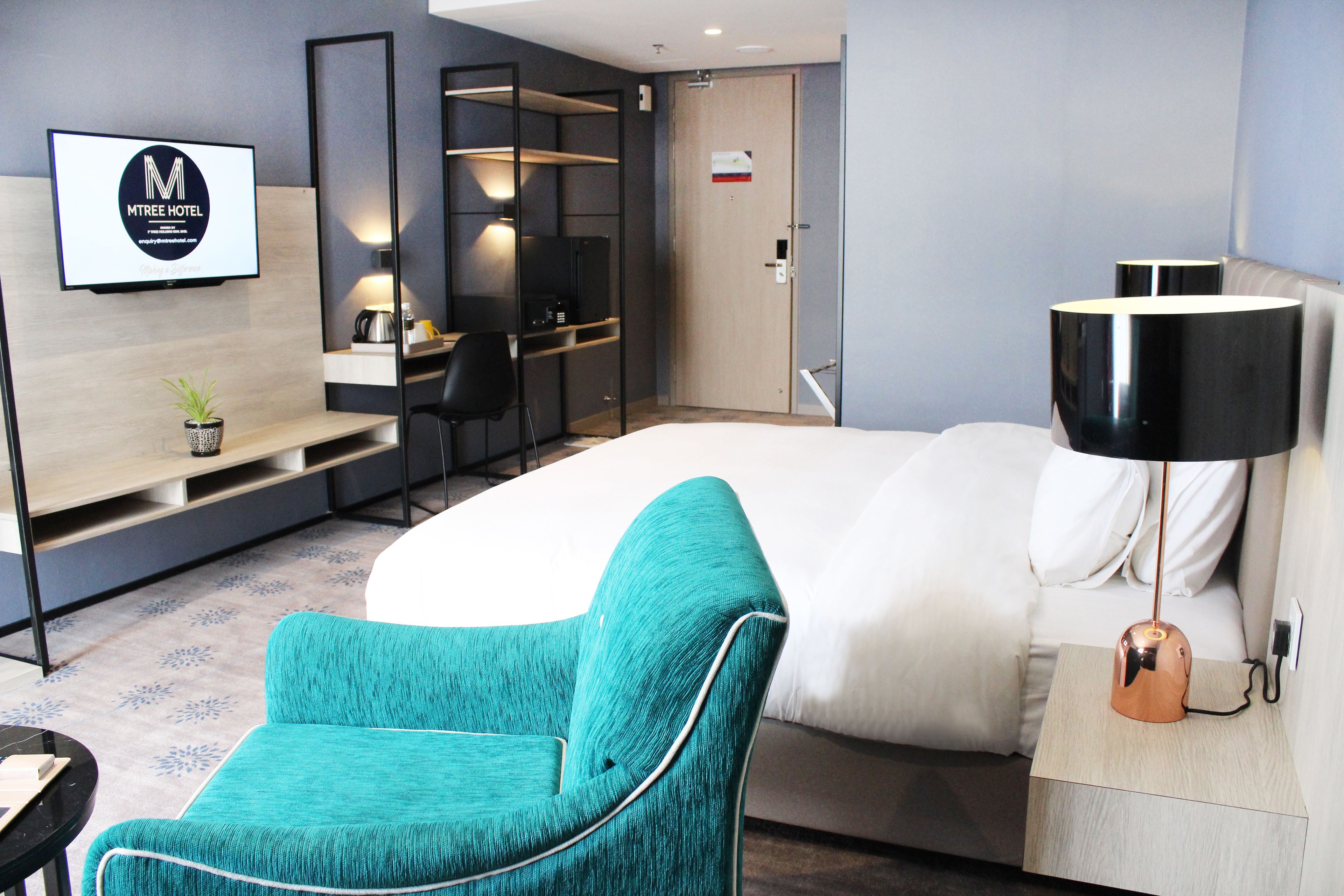 Quarto de hotel moderno com uma cama grande, uma poltrona verde-água, uma escrivaninha com computador e uma TV fixada na parede.