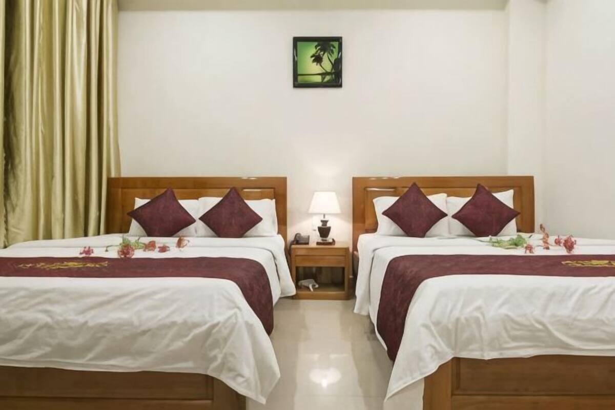 Quarto de hotel com duas camas, cada uma com cabeceira de madeira, roupa de cama branca e almofadas marrons. Há uma mesa de cabeceira com uma luminária e uma pequena mesa entre as camas.