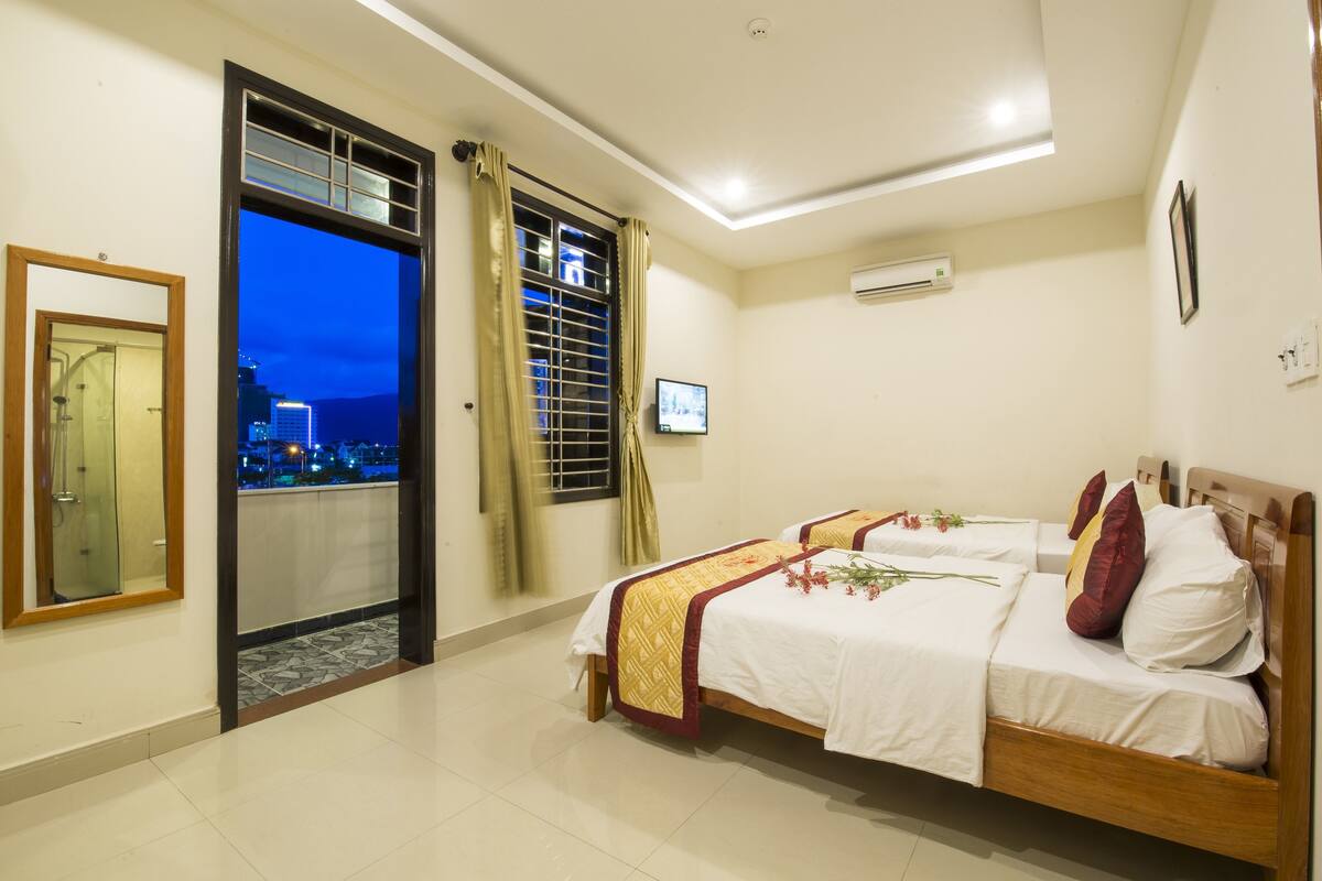 Quarto de hotel com varanda, televisão e cama com roupa de cama branca e almofadas decorativas.