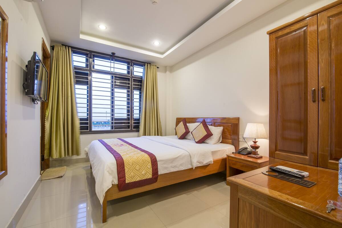 Quarto de hotel com cama, guarda-roupa de madeira, mesa de cabeceira com abajur, televisão na parede e janela com cortinas.