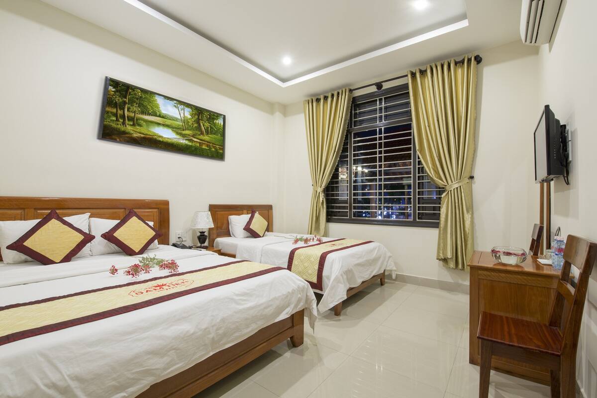 Quarto de hotel com duas camas, uma escrivaninha de madeira, televisão e janela com cortinas.