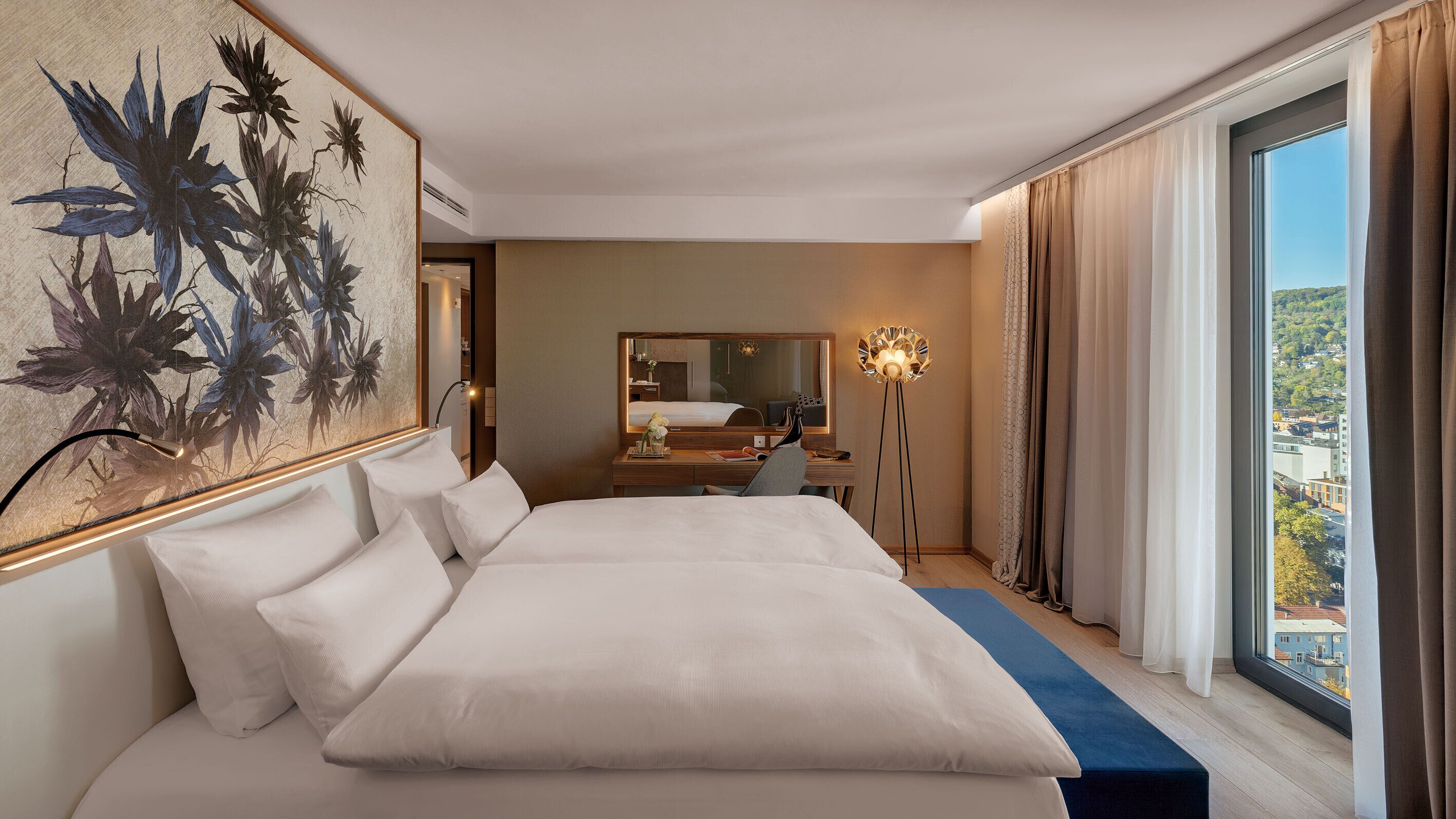 Quarto de hotel com uma cama grande, uma escrivaninha, uma cadeira, um abajur e uma janela ampla com cortinas.