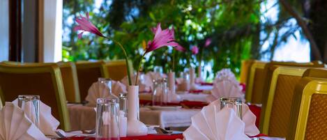 Um restaurante com mesas preparadas para refeições, com toalhas brancas, guardanapos vermelhos e vasos de vidro transparente com flores cor-de-rosa.