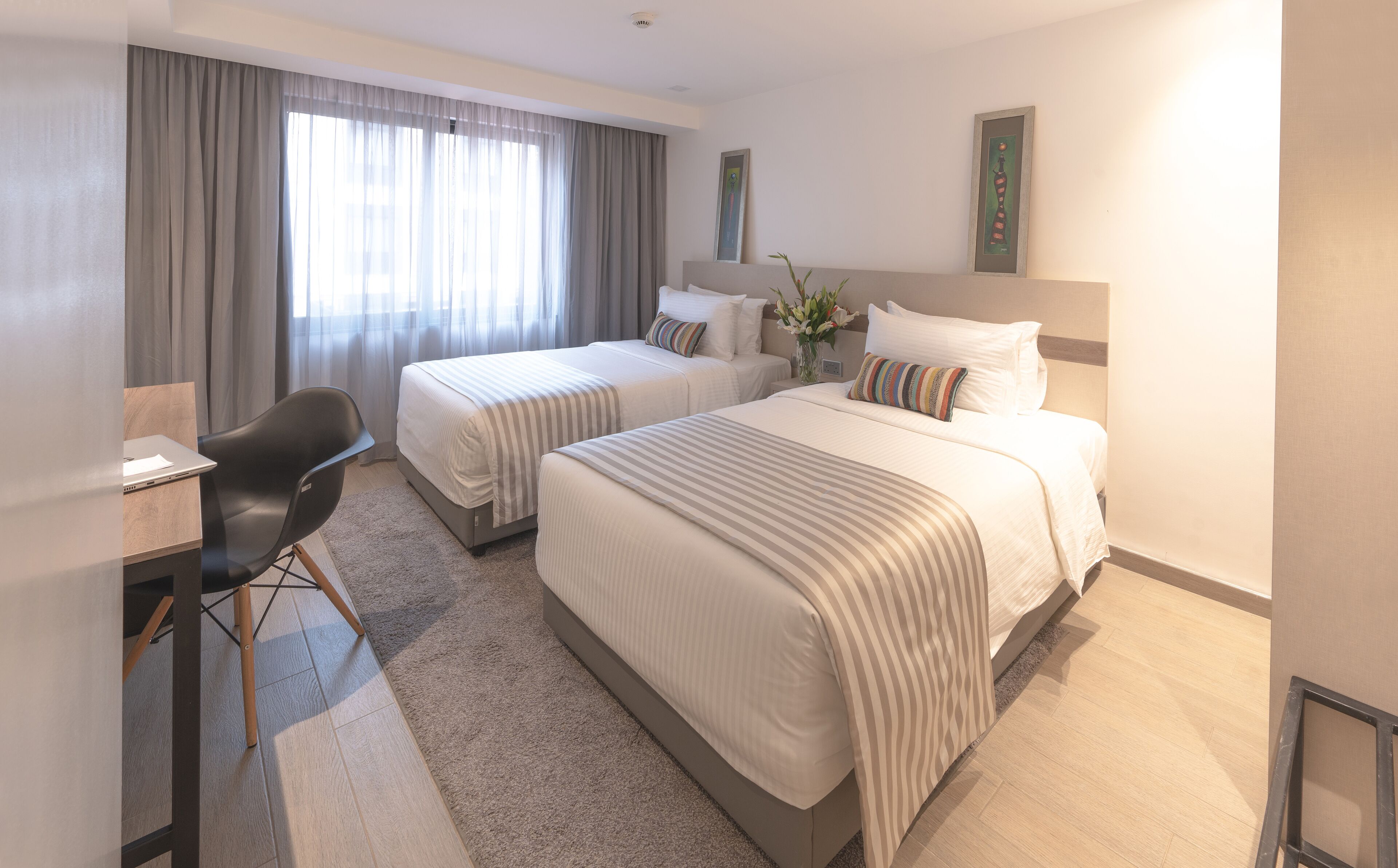 Quarto de hotel com duas camas, uma escrivaninha, uma cadeira e uma janela grande com cortinas.