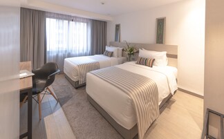 Quarto de hotel com duas camas, uma escrivaninha, uma cadeira e uma janela grande com cortinas.