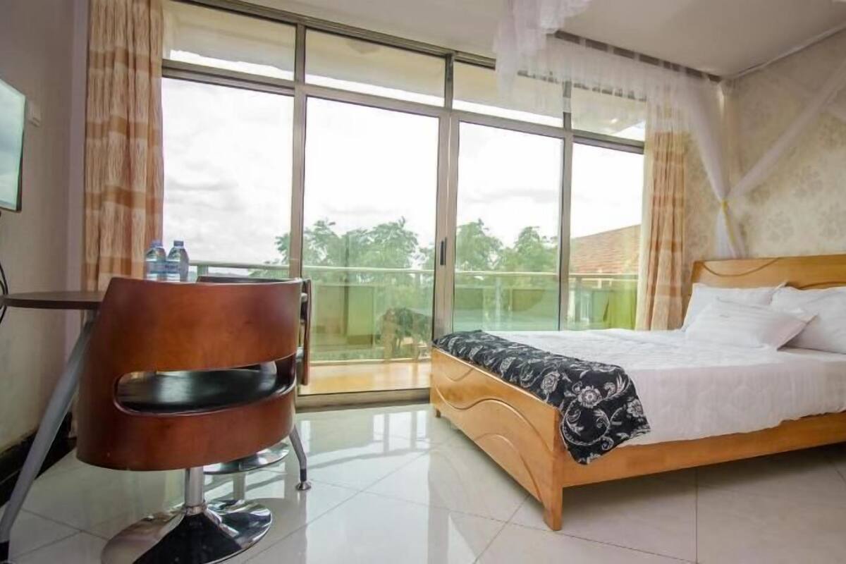 Quarto com cama, poltrona, mesa de cabeceira, telefone e vista através de amplas janelas.