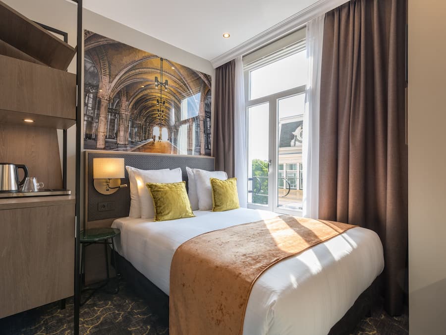 Quarto de hotel com uma cama grande, uma escrivaninha com chaleira, uma mesa pequena e uma luminária na parede.