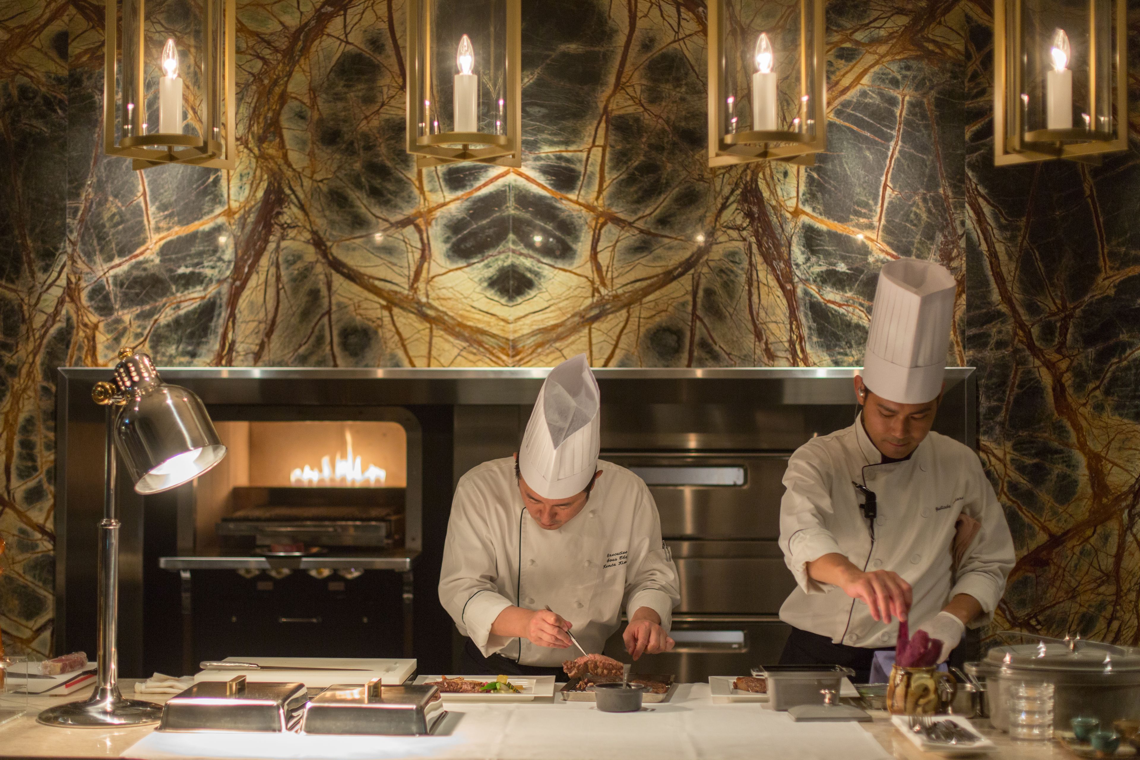 Dois chefs preparando comida em uma cozinha com paredes de mármore e iluminação pendente.