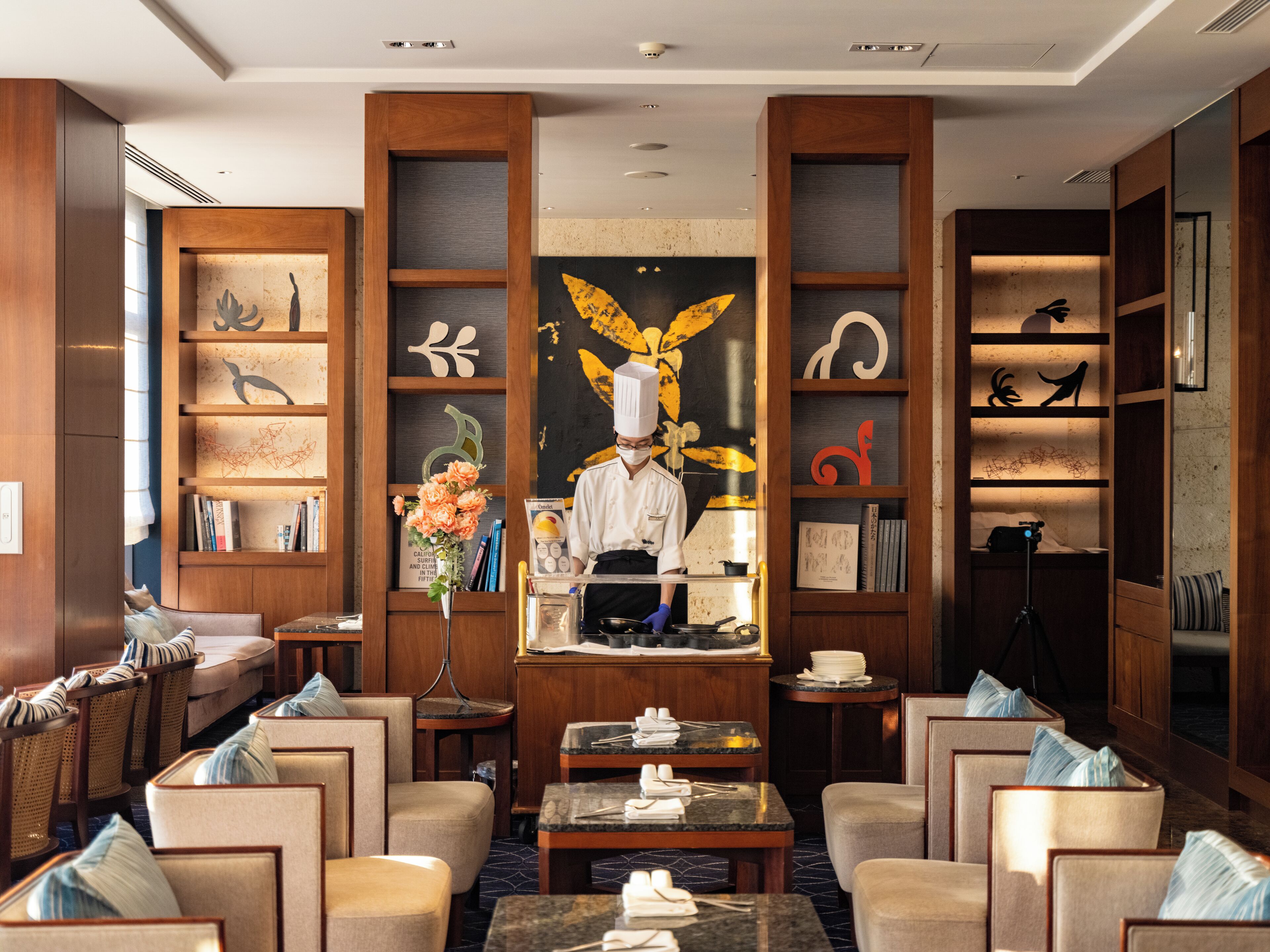Um hall de hotel com uma estátua central de chef, prateleiras de madeira com livros e objetos decorativos, assentos confortáveis e uma mesa de centro de mármore.