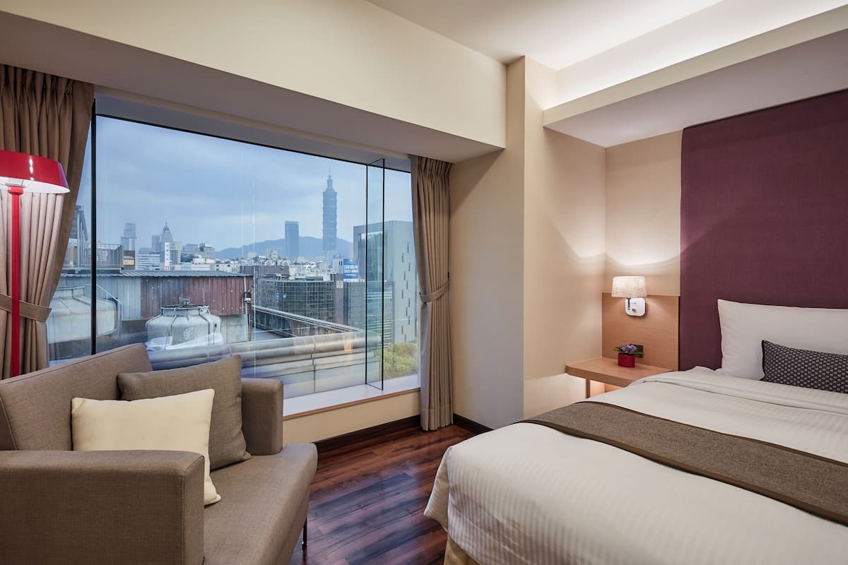 Quarto de hotel com uma janela grande, uma cama, um sofá e uma mesa de cabeceira com abajur.