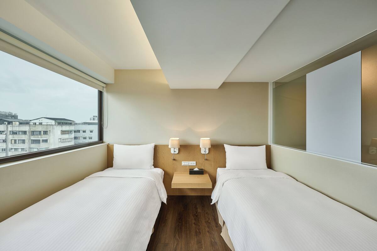 Quarto de hotel com duas camas de solteiro, mesa de cabeceira com abajures, e uma janela grande com vista para a cidade.