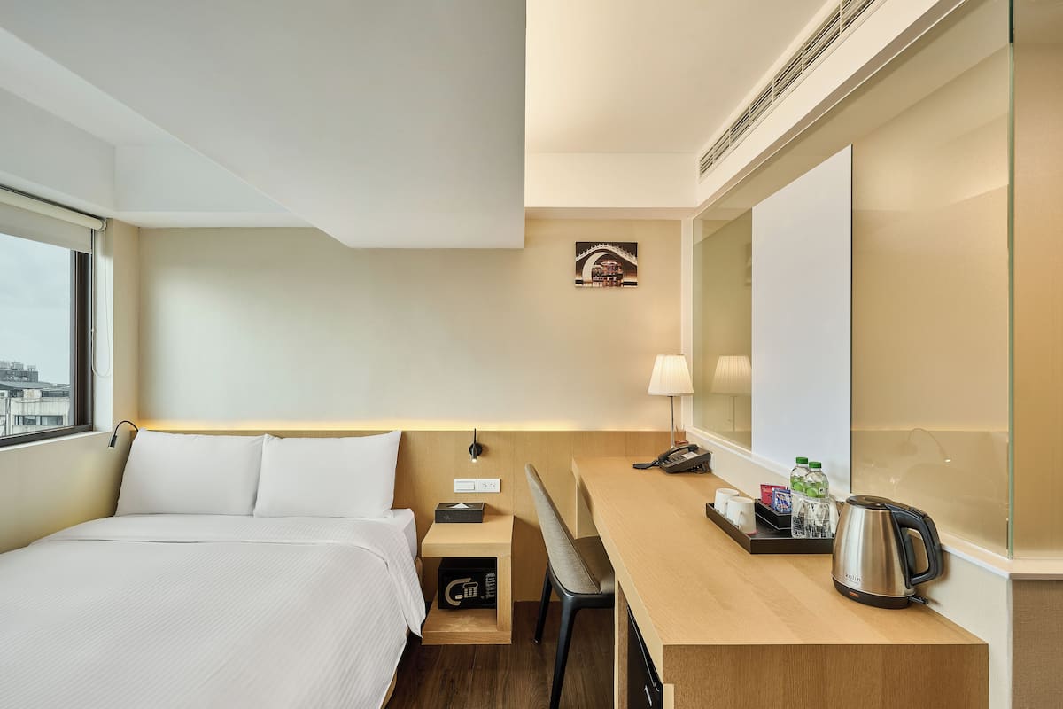 Quarto de hotel com cama, escrivaninha, cadeira, luminária, chaleira e uma pequena mesa com artigos de higiene pessoal.