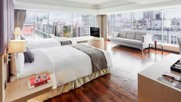 Quarto de hotel moderno com uma cama grande, um sofá, uma TV e uma escrivaninha. O quarto possui janelas amplas com vista para a cidade.