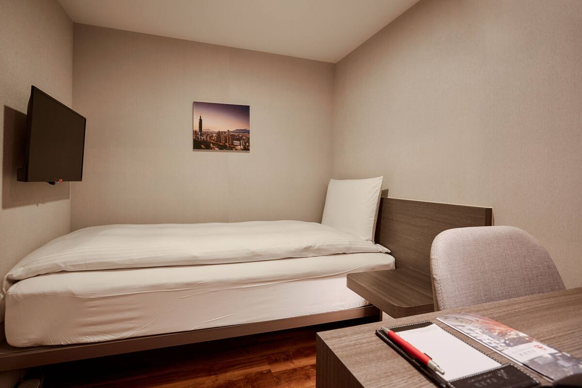 Quarto de hotel com cama, escrivaninha, cadeira, televisão de tela plana e uma fotografia da paisagem urbana na parede.