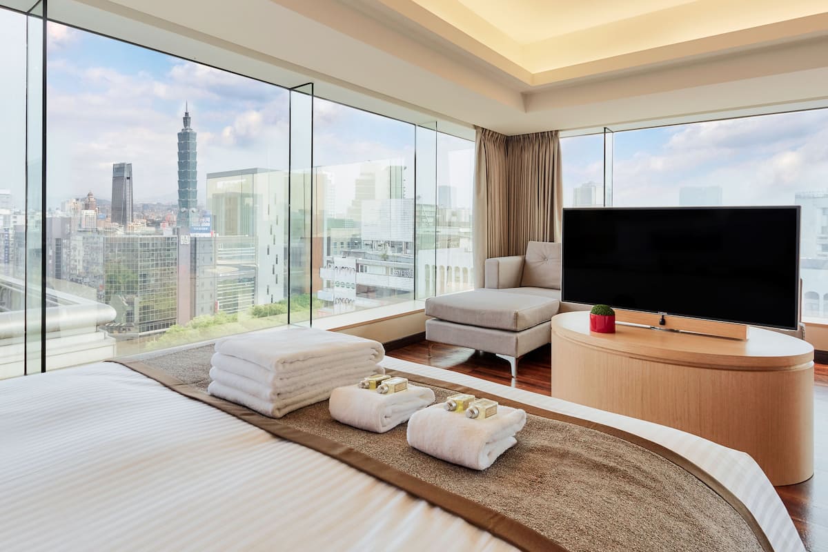 Quarto de hotel moderno com uma cama grande, TV de tela plana, área de estar e vista para a cidade.