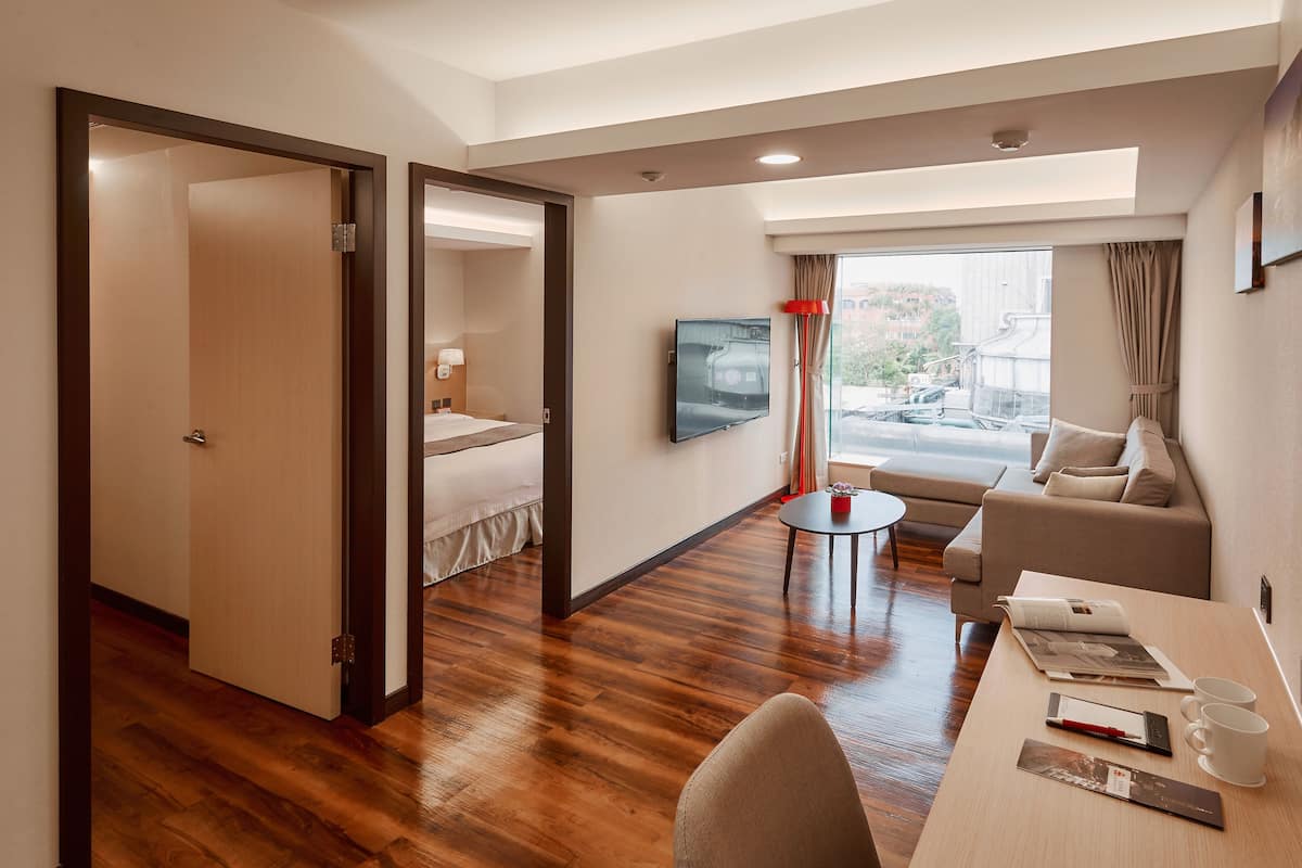 Quarto de hotel com piso de madeira, uma cama, televisão, sofá, mesa de jantar e janela com vista para o exterior.