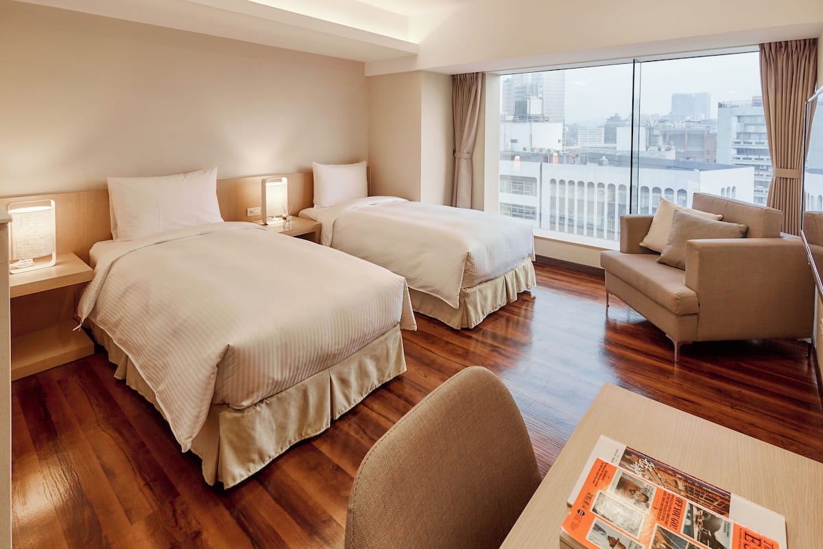 Quarto de hotel com duas camas, um sofá, uma mesa e vista para a cidade.