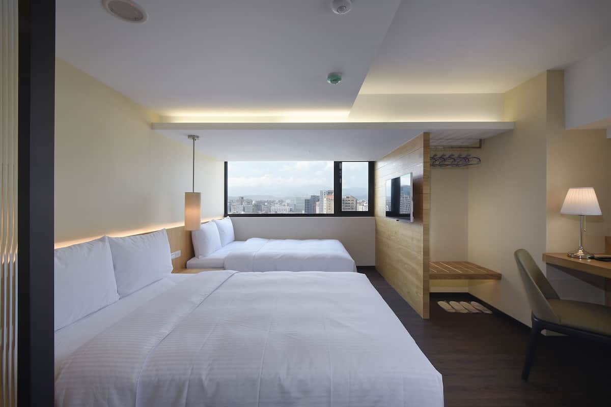 Quarto de hotel com duas camas, uma escrivaninha, uma cadeira e uma janela grande com vista para a cidade.