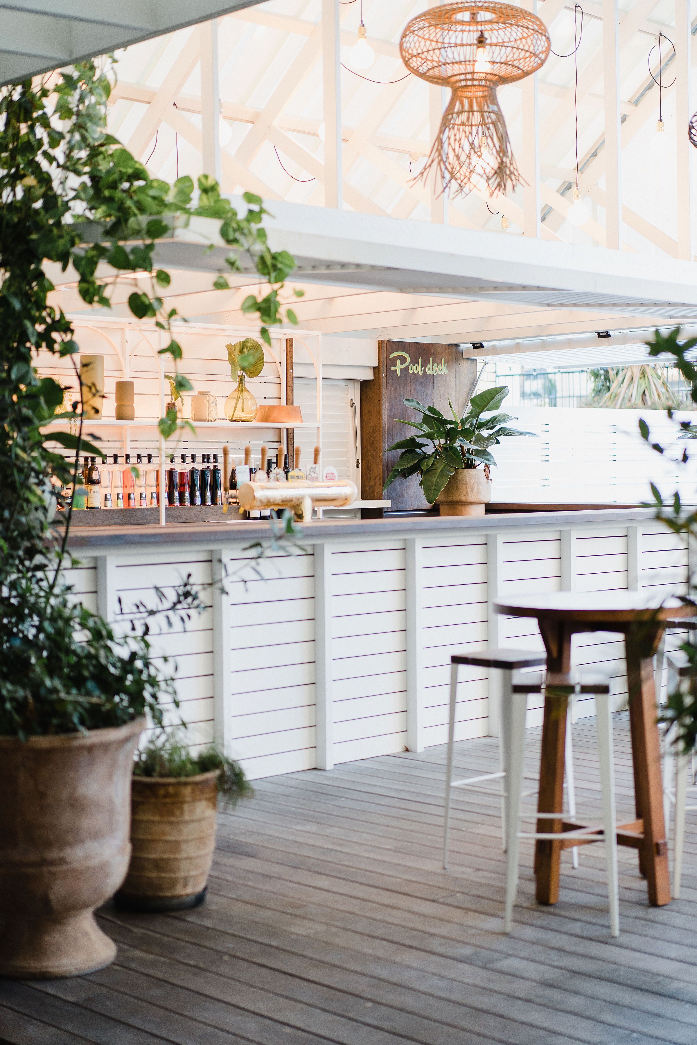 Uma área de bar com placa de deck de piscina, balcão de madeira, plantas em vasos e luminárias pendentes.