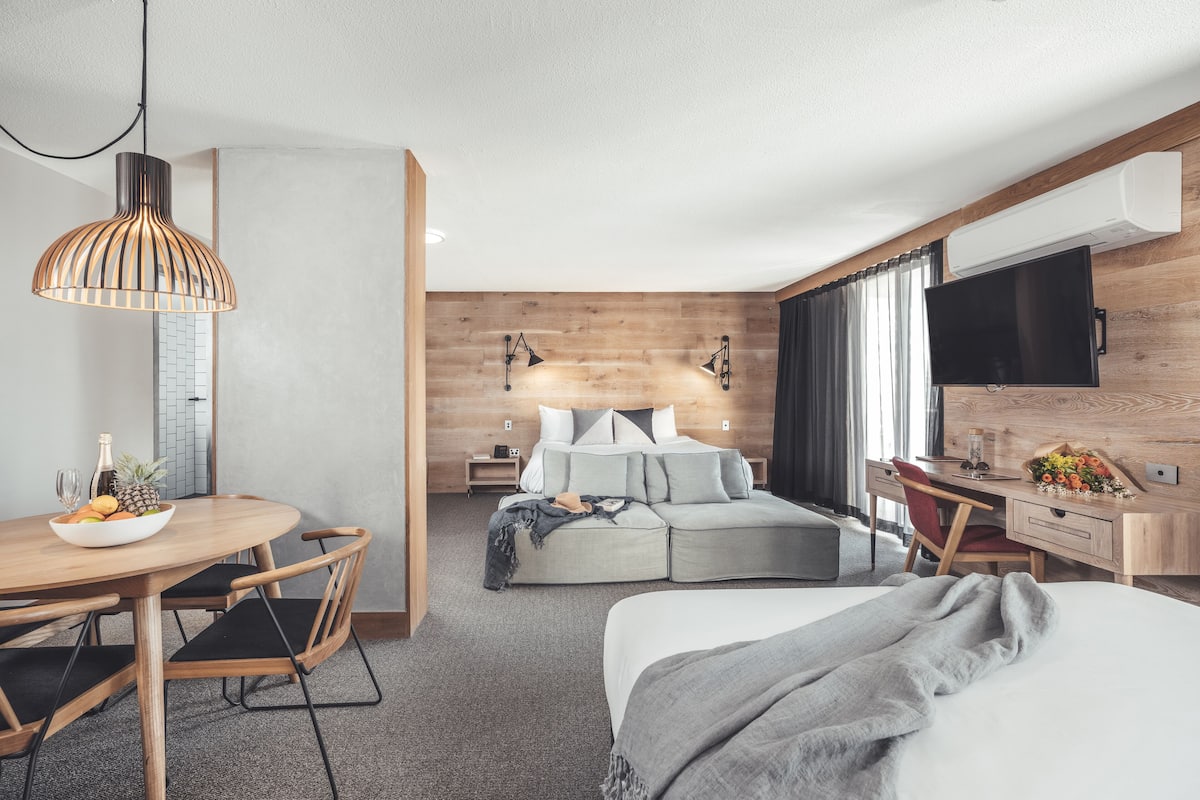 Quarto de hotel moderno com cabeceira de madeira, televisão de tela plana, área de refeições com mesa e cadeiras de madeira, e cama aconchegante com manta cinza.