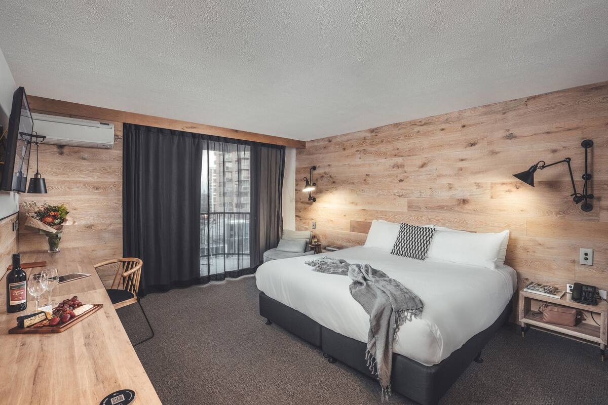 Quarto de hotel com uma cama grande, uma escrivaninha com uma garrafa de vinho e copos, uma cadeira, uma mesa de cabeceira com abajur e vista para edifícios pela janela.