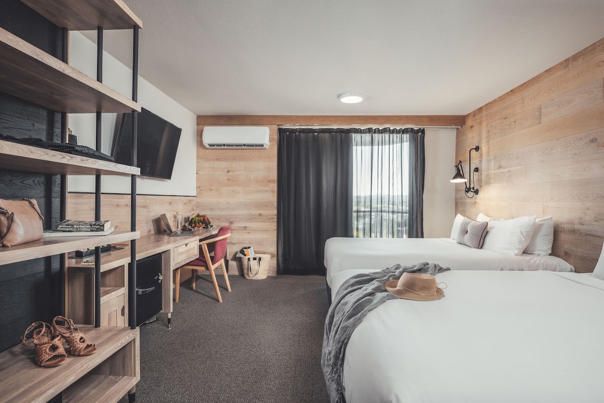 Quarto de hotel com duas camas, uma mesa de trabalho com cadeira, televisão e janela com cortinas.