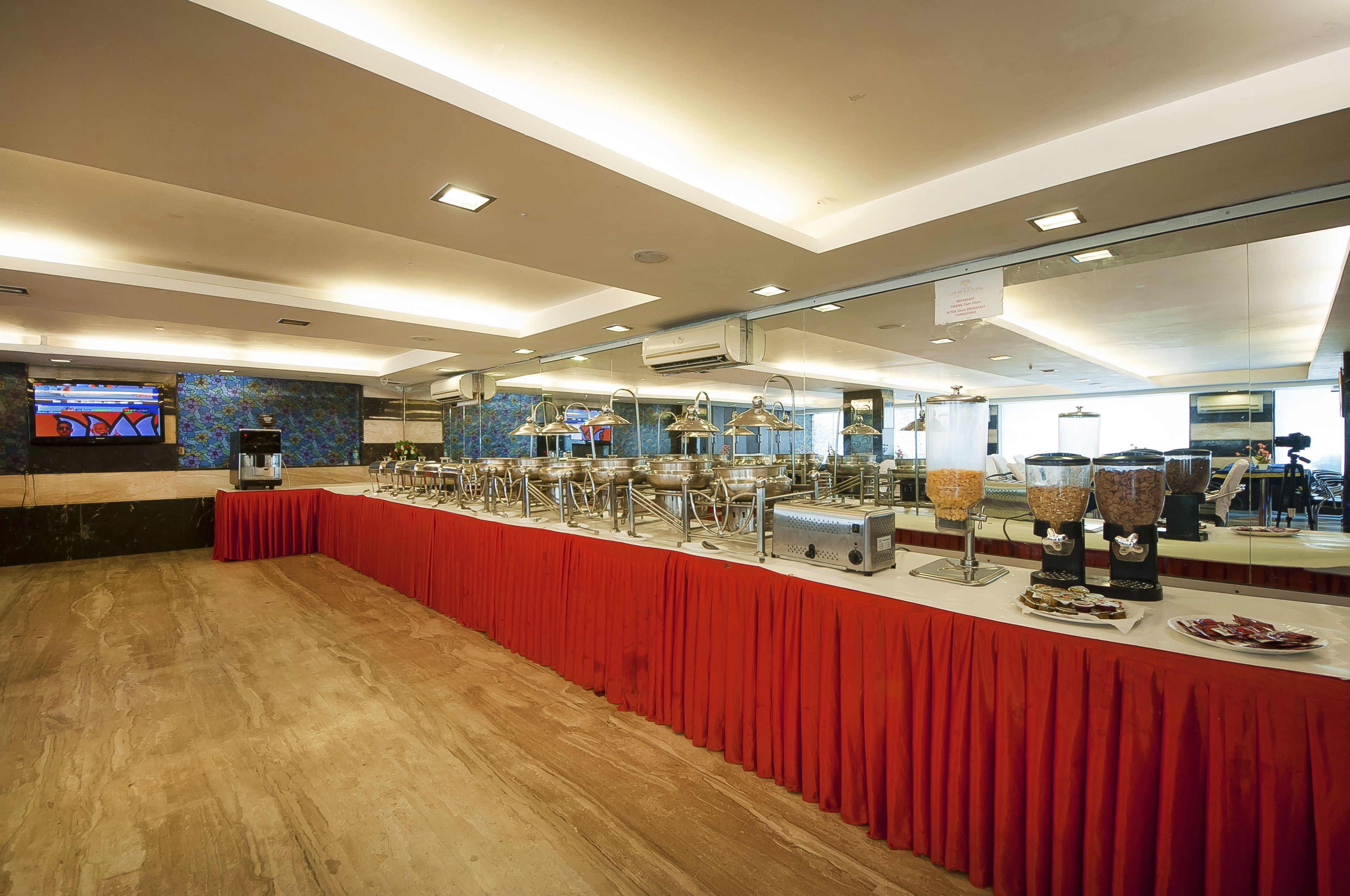 Disposição de buffet com uma longa mesa coberta por toalha vermelha, diversos dispensadores de comida e uma tela de televisão na parede.