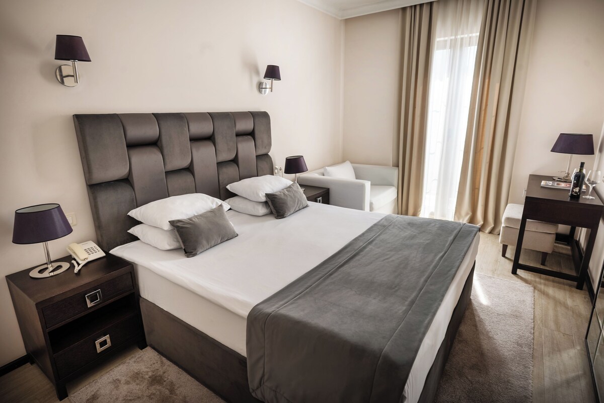 Quarto de hotel com uma cama grande, mesas de cabeceira, uma escrivaninha e uma cadeira.
