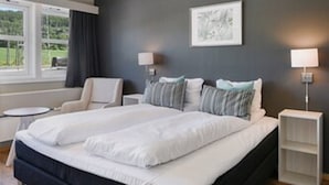 Quarto de hotel moderno com uma cama grande, mesas de cabeceira, uma cadeira e uma janela com vista para vegetação.