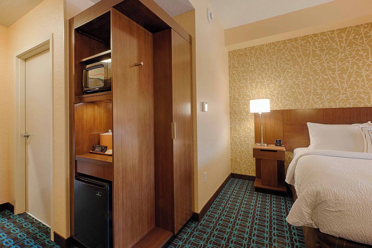 Quarto de hotel com cama, guarda-roupa, pequeno frigorífico, mesa de cabeceira com luminária e carpete estampado.