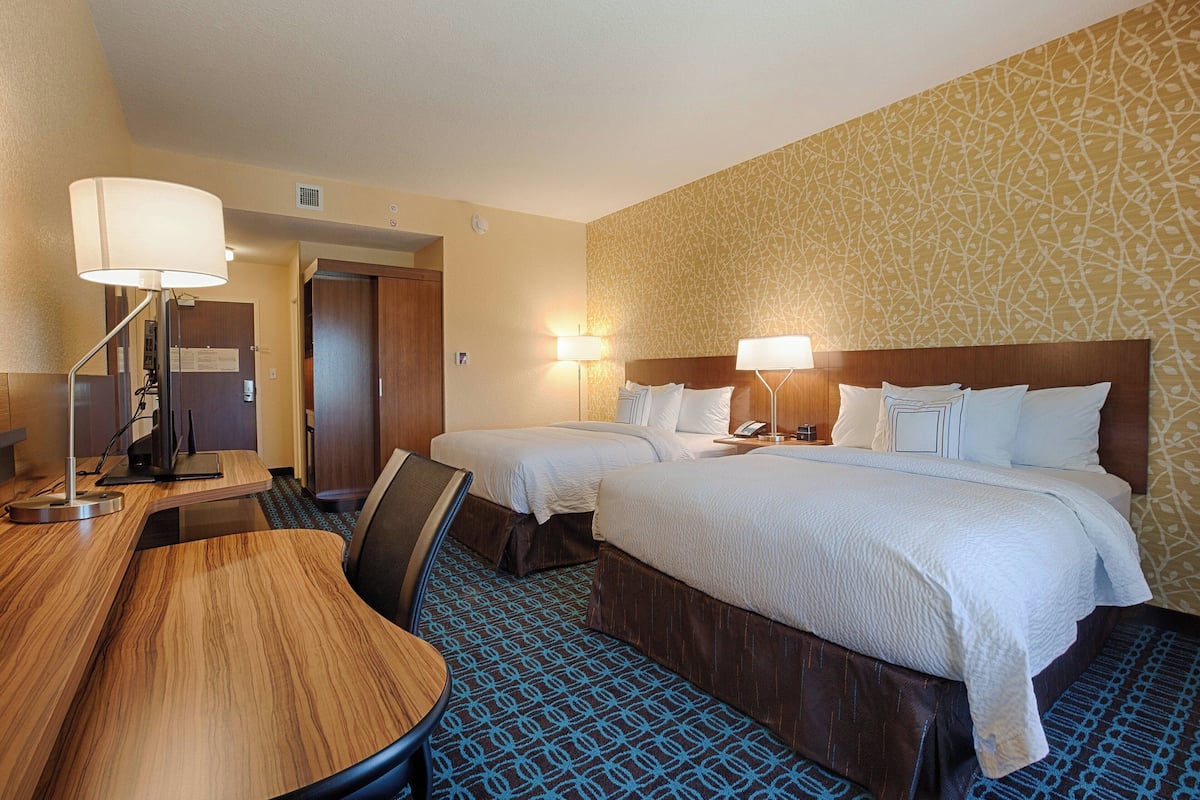 Quarto de hotel com duas camas, uma escrivaninha e uma cadeira. Há duas luminárias de cabeceira e um telefone na escrivaninha.