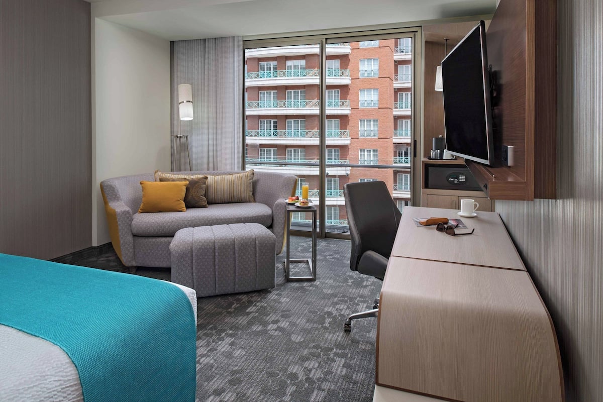 Quarto de hotel com sofá, uma pequena mesa com bebida, escrivaninha e televisão fixada na parede.