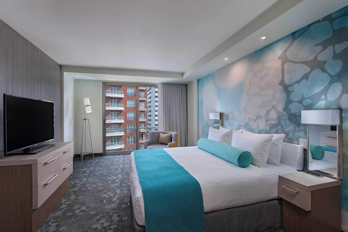Quarto de hotel com uma cama grande, televisão, escrivaninha e vista para um edifício pela janela.