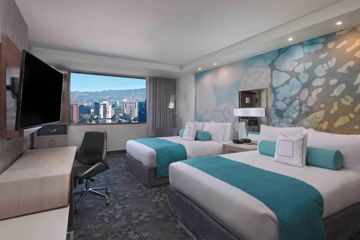 Quarto de hotel com duas camas, uma escrivaninha, uma cadeira, uma televisão e vista para a cidade.