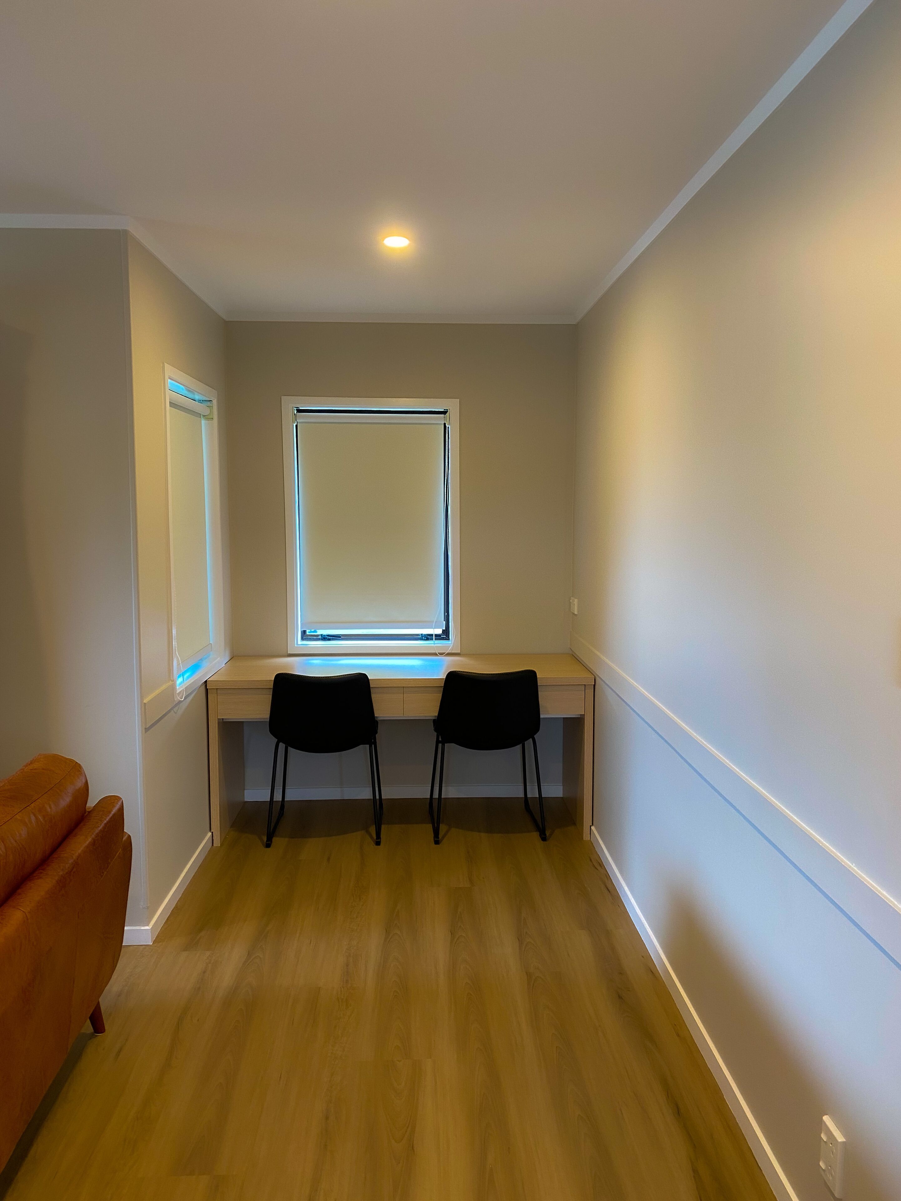 Um quarto pequeno e bem iluminado, com uma escrivaninha, duas cadeiras, uma janela e um sofá de couro marrom.