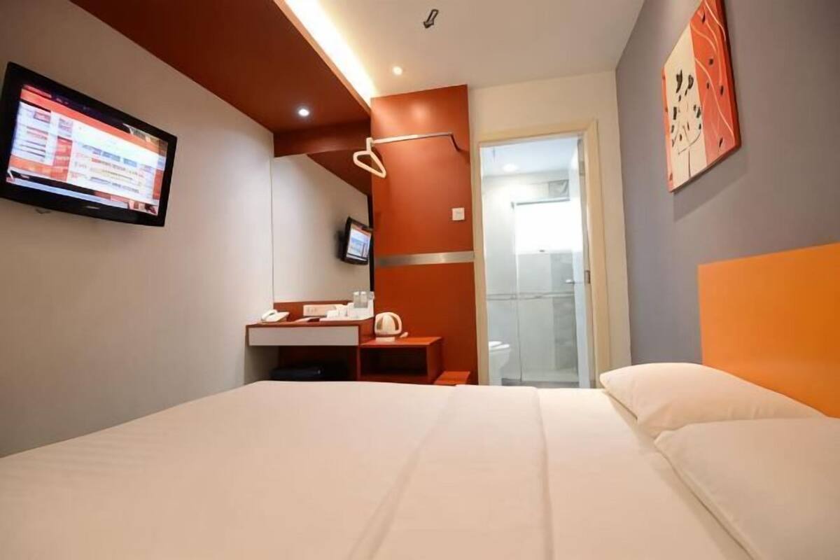 Quarto de hotel com cama, escrivaninha com computador, televisão e banheiro visível por uma porta aberta.