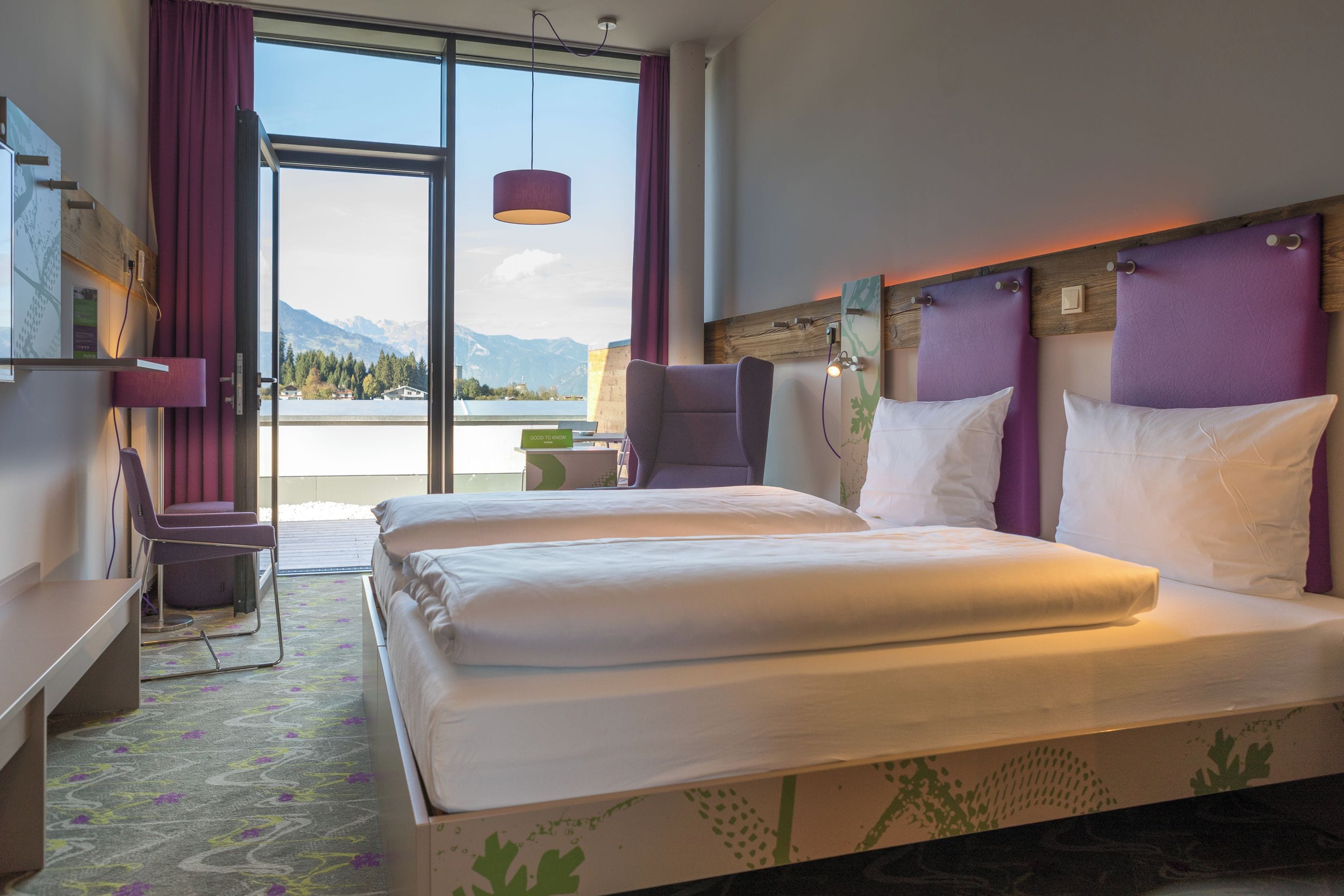 Quarto de hotel com cama, escrivaninha, cadeira e vista para montanhas pela janela.