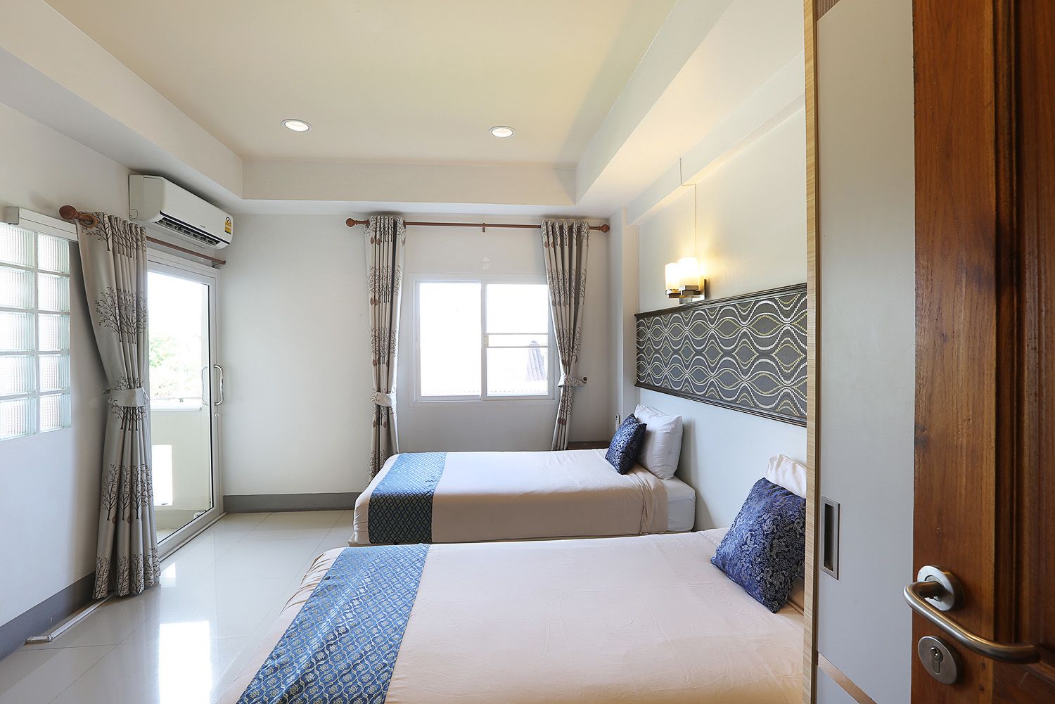 Quarto de hotel com duas camas, uma janela grande, um aparelho de ar condicionado e um painel decorativo na parede.