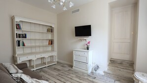 Sala de estar com estante branca, televisão de tela plana, mesa de cabeceira branca, sofá cinza e piso de madeira.