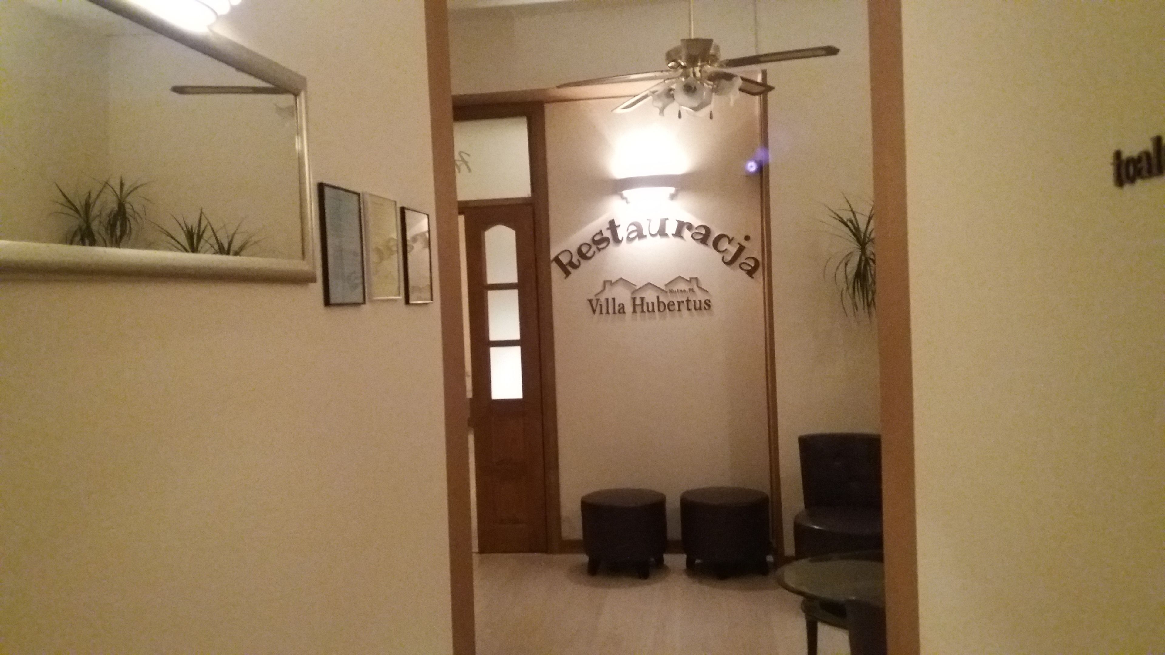 Corredor que leva a um restaurante chamado 'Restaurante Villa Hubertus', com quadros nas paredes, um ventilador de teto e dois banquinhos em primeiro plano.
