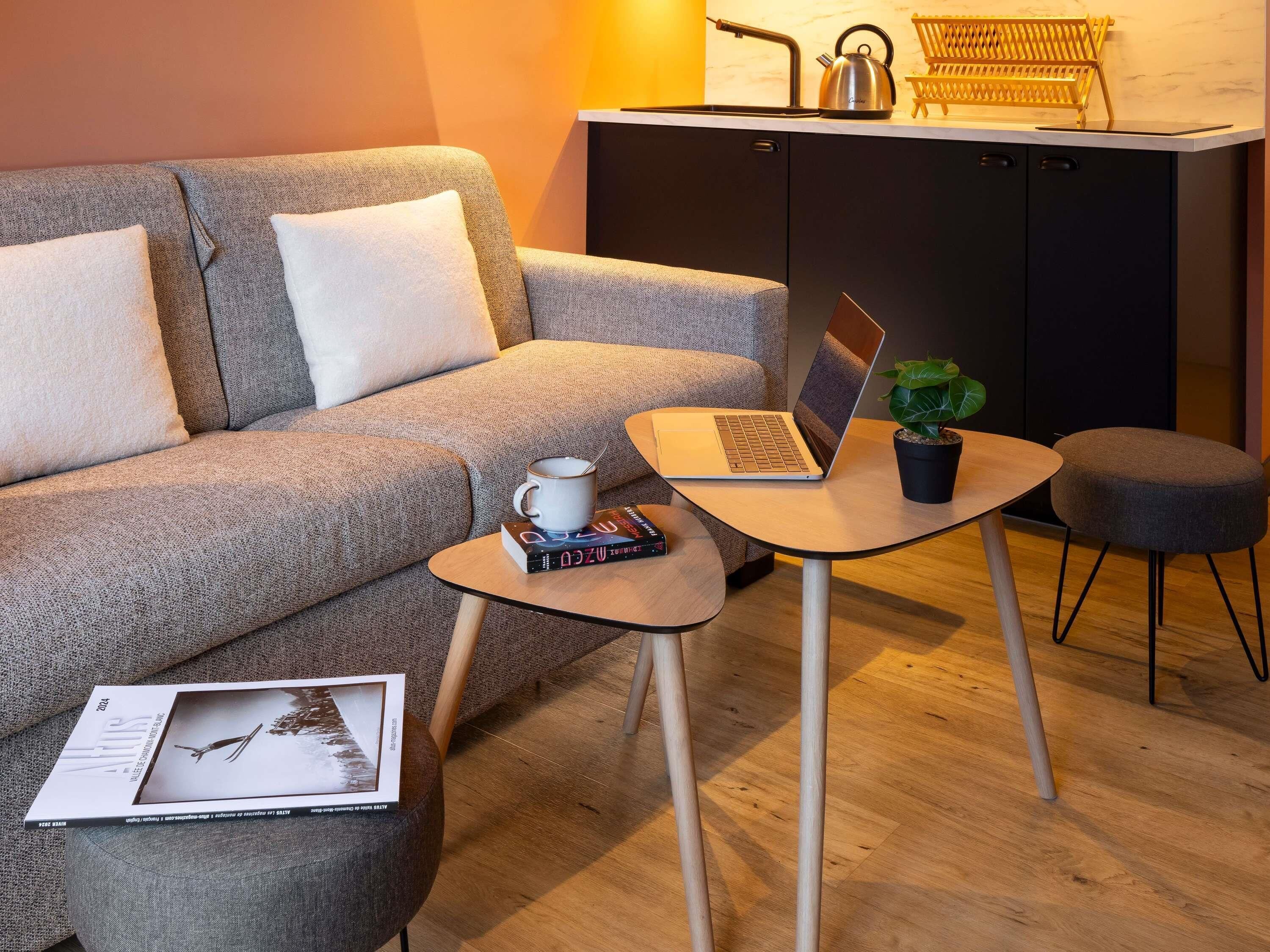 Uma sala de estar moderna com um sofá cinzento, uma pequena mesa de madeira com um laptop, uma caneca branca e uma planta em vaso.