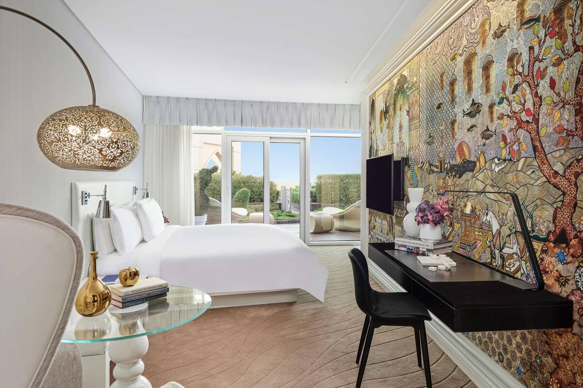 Quarto de hotel com cama grande, mesa de vidro, cadeira, televisão e varanda com vista para o verde.