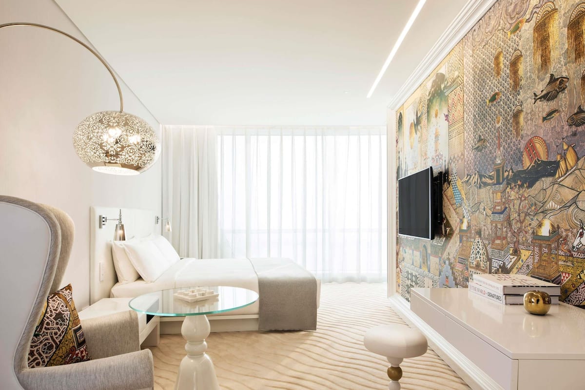 Quarto de hotel moderno com cama, mesa de centro de vidro, área de estar com uma cadeira, pequena mesa com um livro e parede com um mural decorativo.