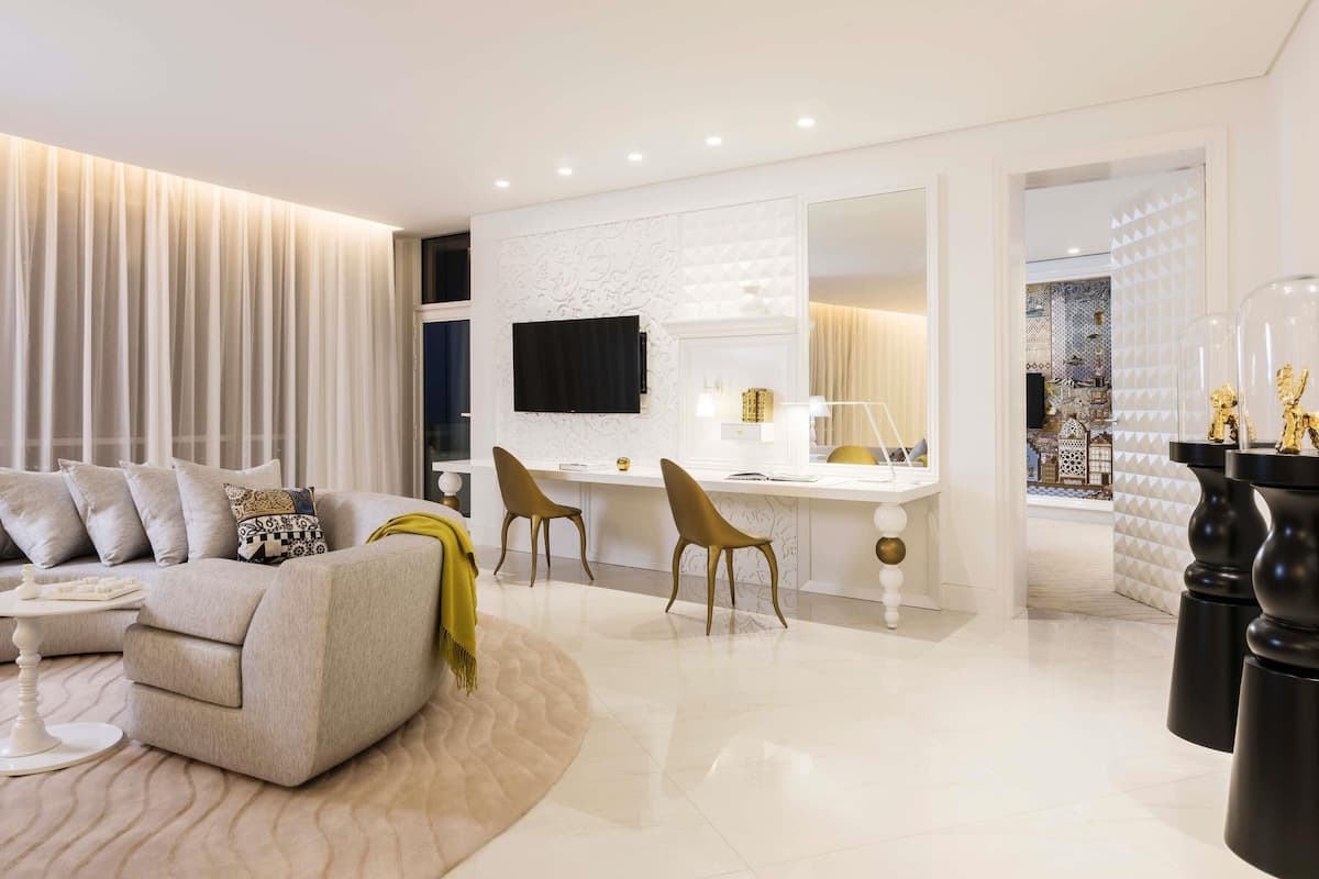Uma sala de estar moderna com um sofá, uma área de refeições com cadeiras, uma TV fixada na parede e um espelho decorativo.