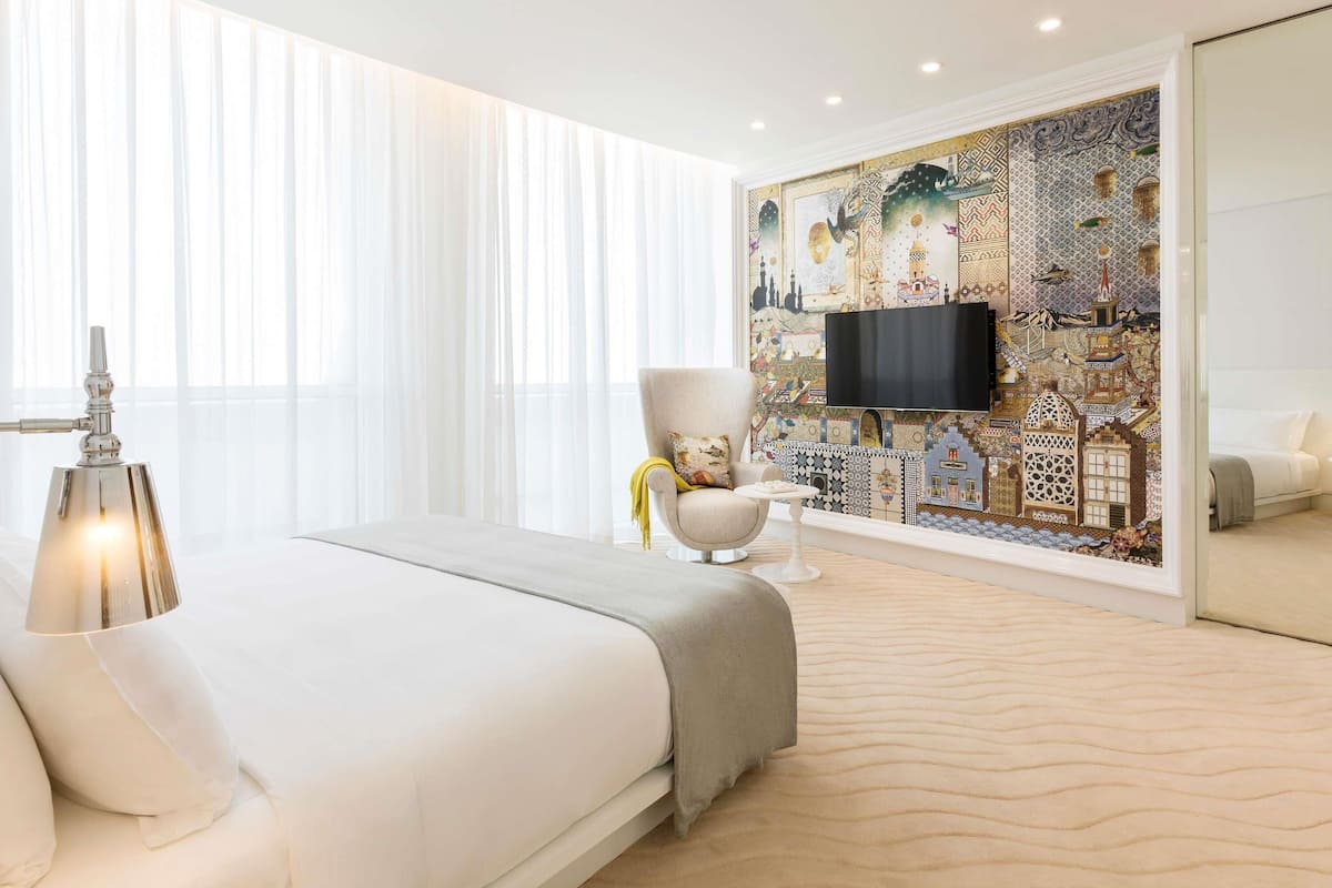 Quarto de hotel moderno com uma cama grande, uma TV de tela plana fixada em uma parede colorida e uma janela ampla com cortinas translúcidas.
