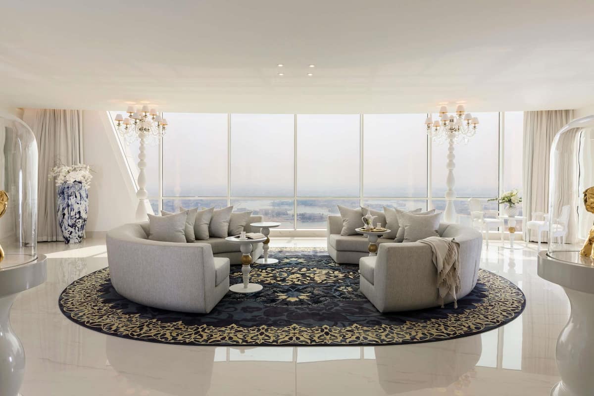 Uma sala de estar ampla, com uma grande janela que oferece vista para a cidade, uma mesa de centro redonda e um candelabro.