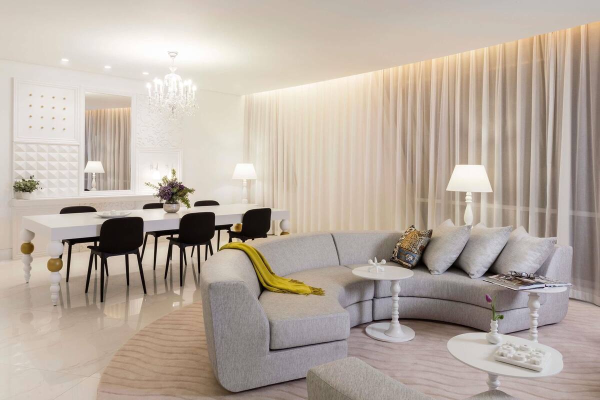 Uma sala de estar moderna com área de jantar, um candelabro e uma janela grande com cortinas.