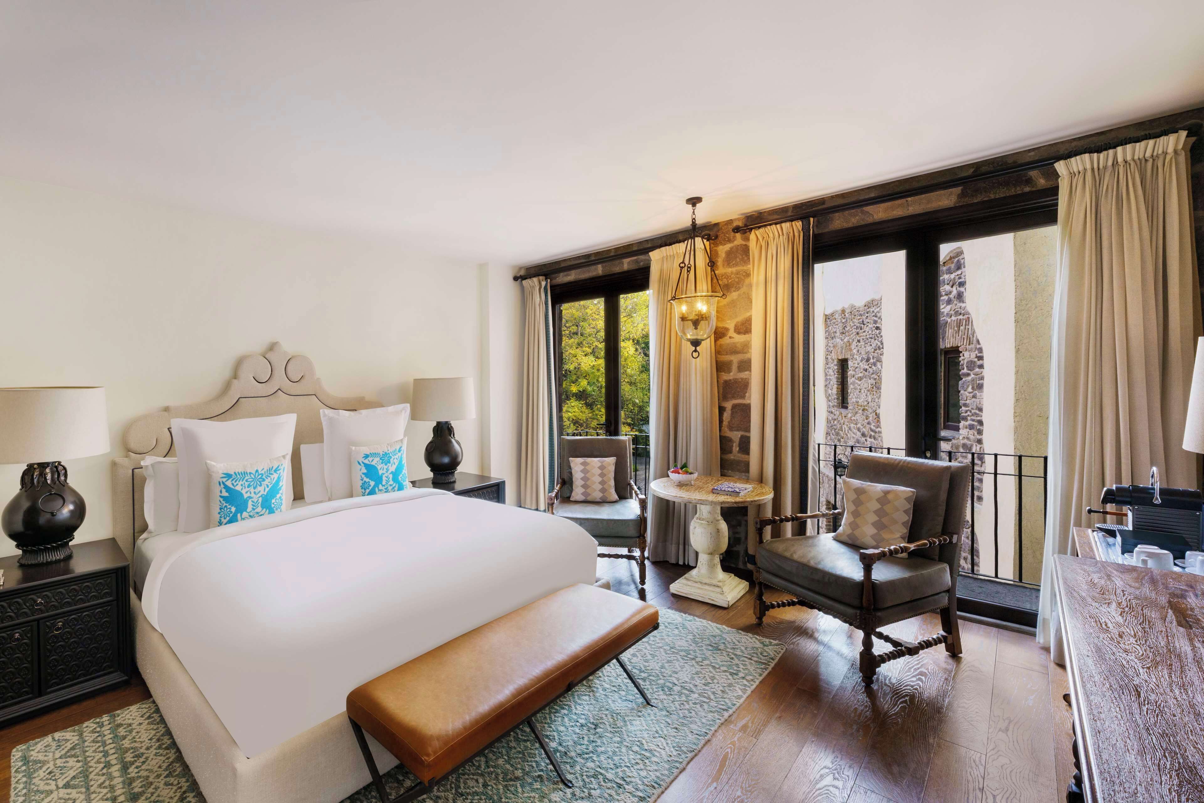 Quarto de hotel com uma cama grande, um banco, uma mesinha, uma cadeira e vista para o exterior.