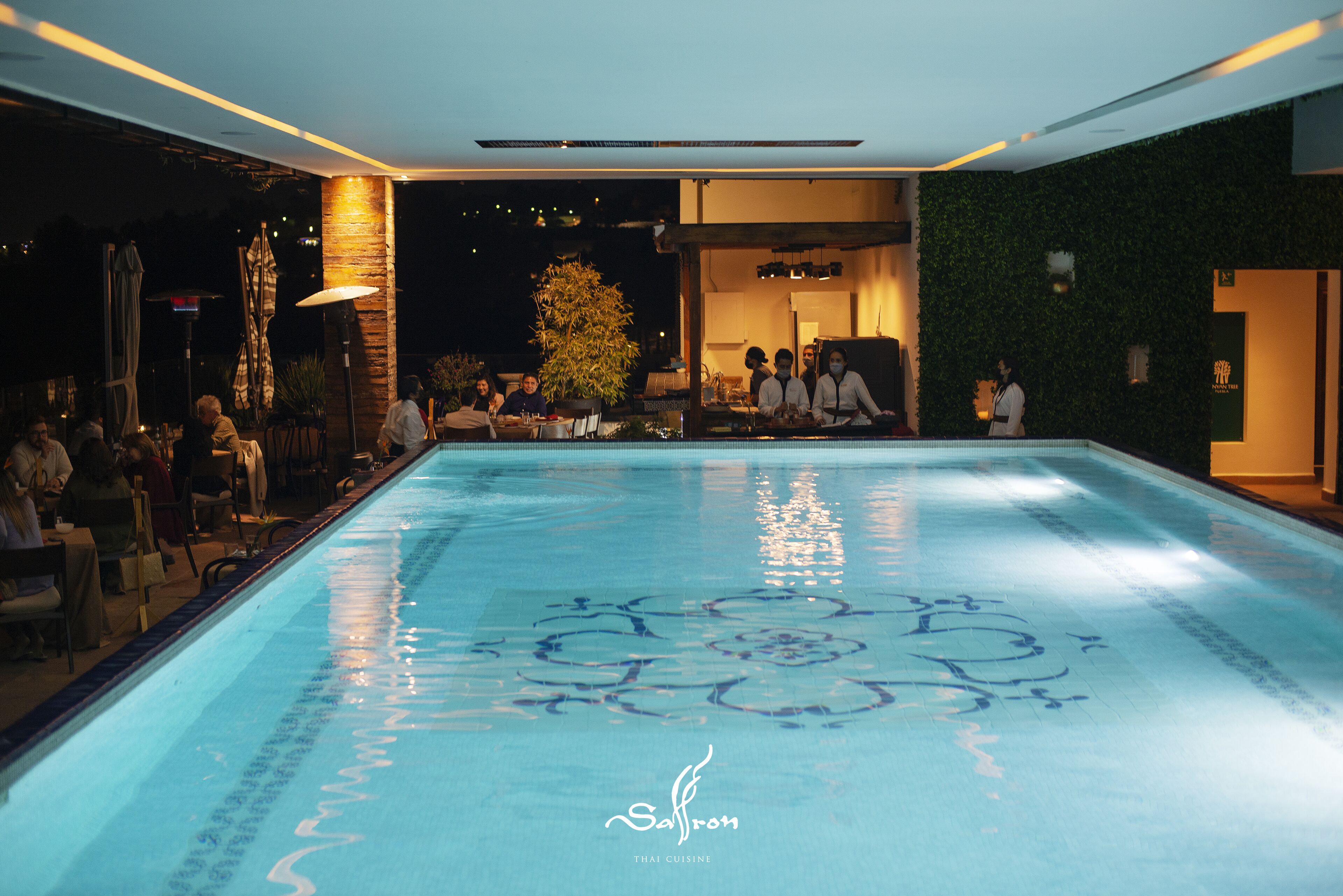 Uma piscina com um desenho decorativo, rodeada por mesas e cadeiras, e um grupo de pessoas reunidas em torno de uma mesa.
