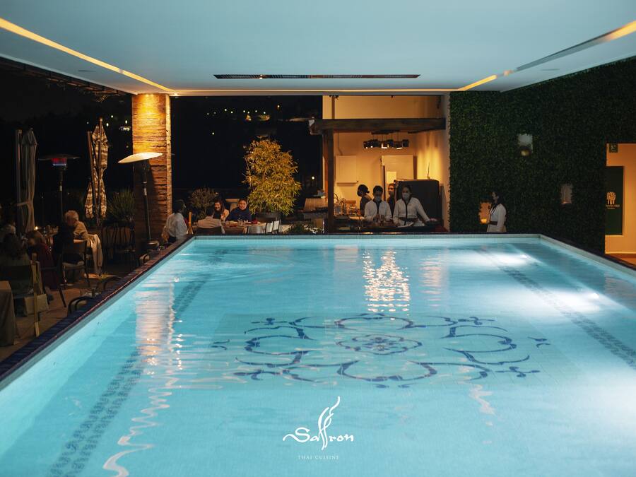Uma piscina com um desenho decorativo, rodeada por mesas e cadeiras, e um grupo de pessoas reunidas em torno de uma mesa.